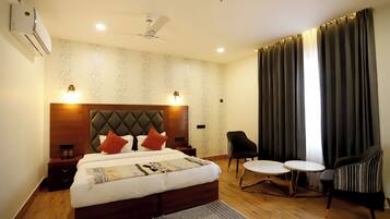 Quarto de hotel moderno com uma cama grande, um ventilador de teto, área de estar com duas cadeiras e uma mesa pequena, e uma janela grande com cortinas.