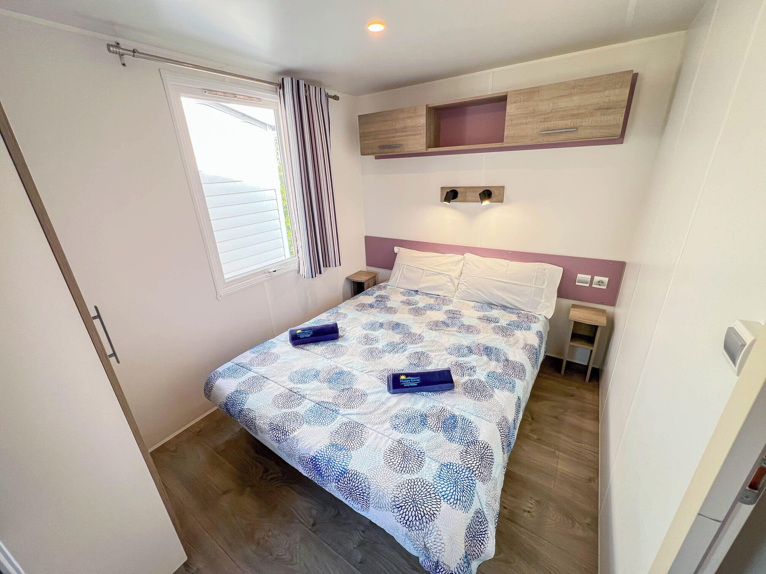 Quarto com cama, janela com cortinas, mesa de cabeceira e prateleira fixada na parede.