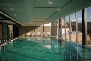Piscina interior moderna com água cristalina, iluminada por grandes janelas, e vista para uma paisagem montanhosa.