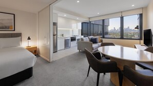 Quarto de hotel moderno com cama, mesa de jantar e sofá. Possui uma cozinha compacta com geladeira e pia. O quarto tem grandes janelas com vista para a cidade.