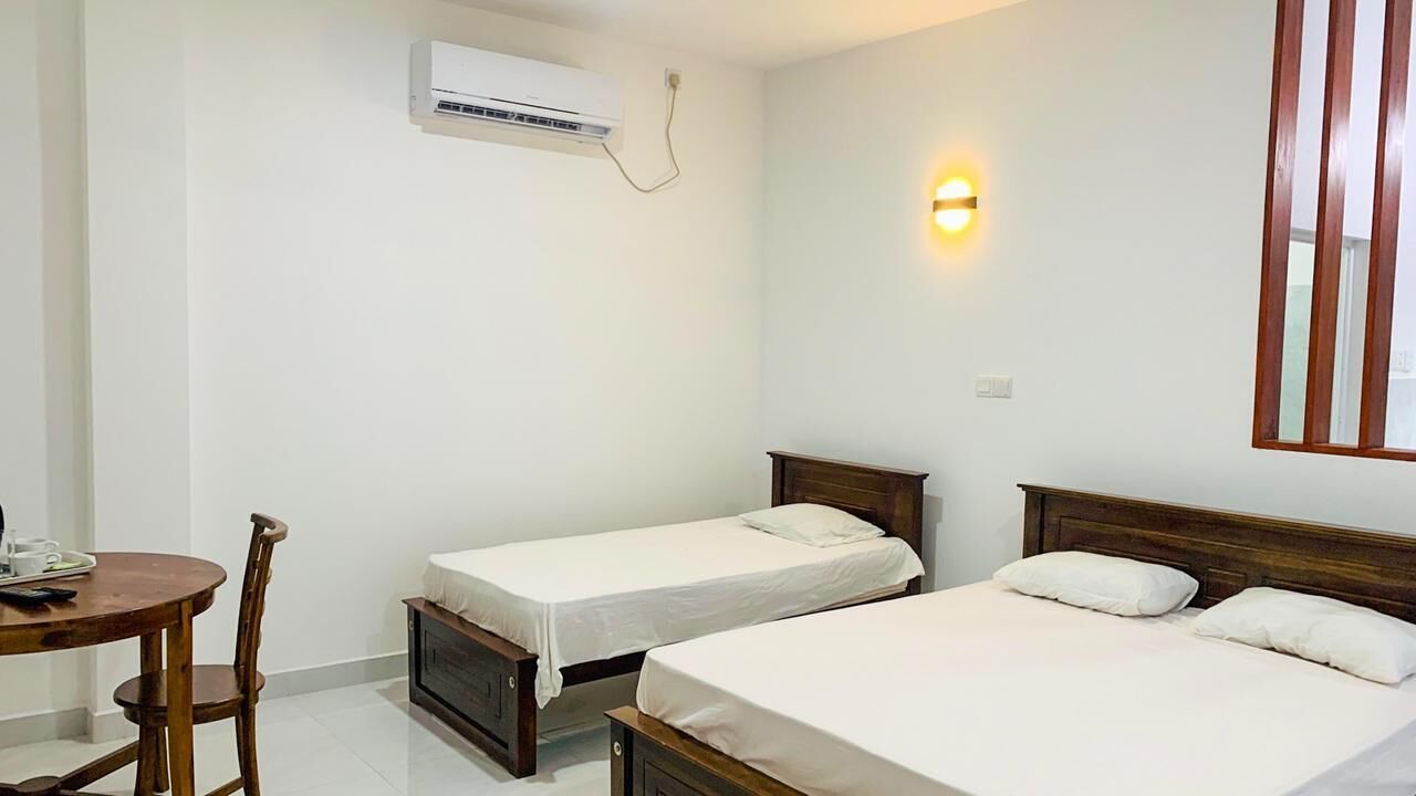 Quarto de hotel com duas camas de solteiro, uma mesa de madeira com cadeiras, uma pequena escrivaninha, unidade de ar condicionado e luminária fixada na parede.
