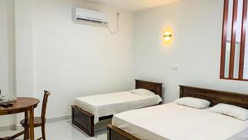 Quarto de hotel com duas camas de solteiro, uma mesa de madeira com cadeiras, uma pequena escrivaninha, unidade de ar condicionado e luminária fixada na parede.