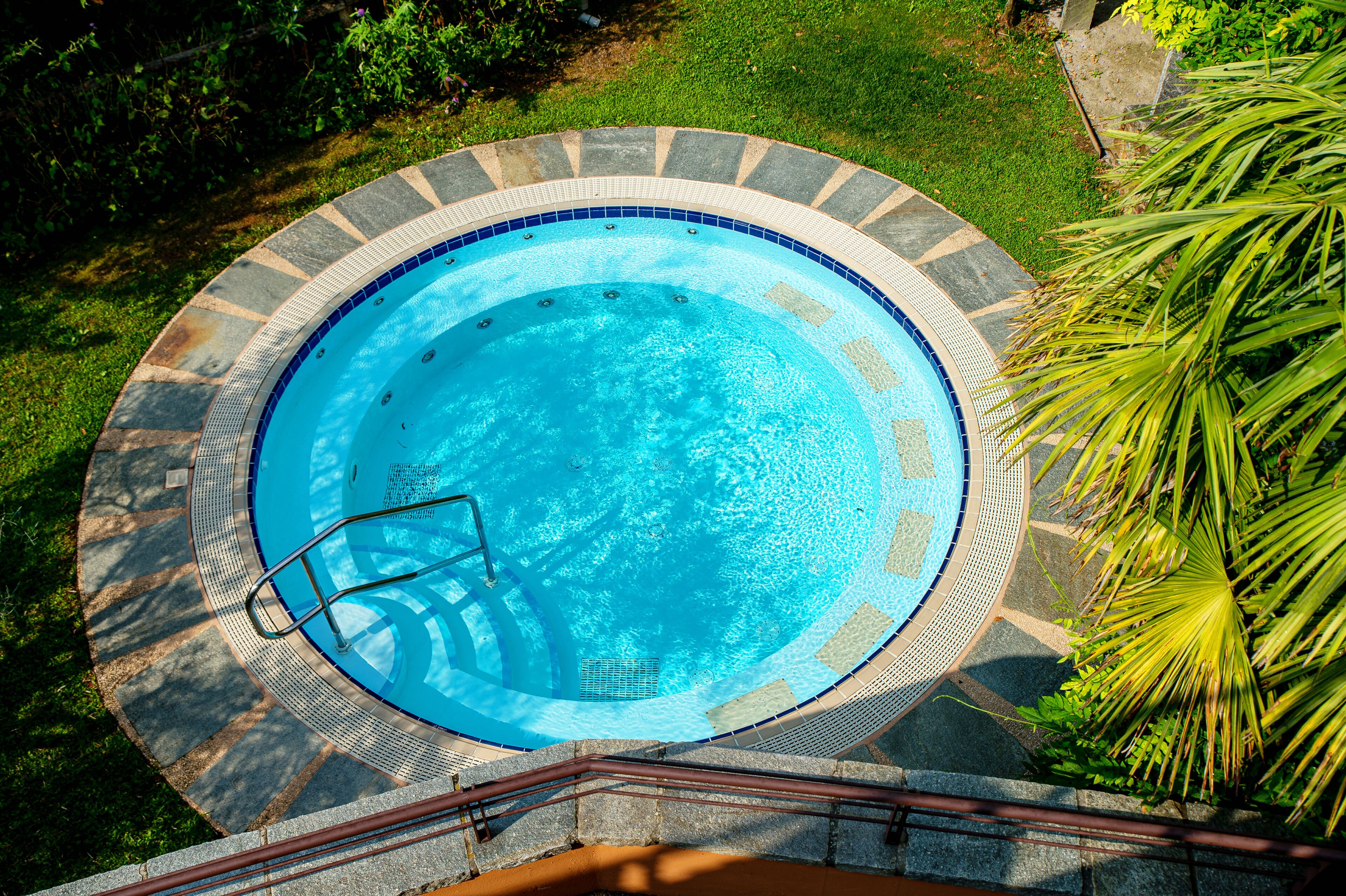 Piscina circular com borda azulejada, rodeada por um deck de pedra e uma área gramada.