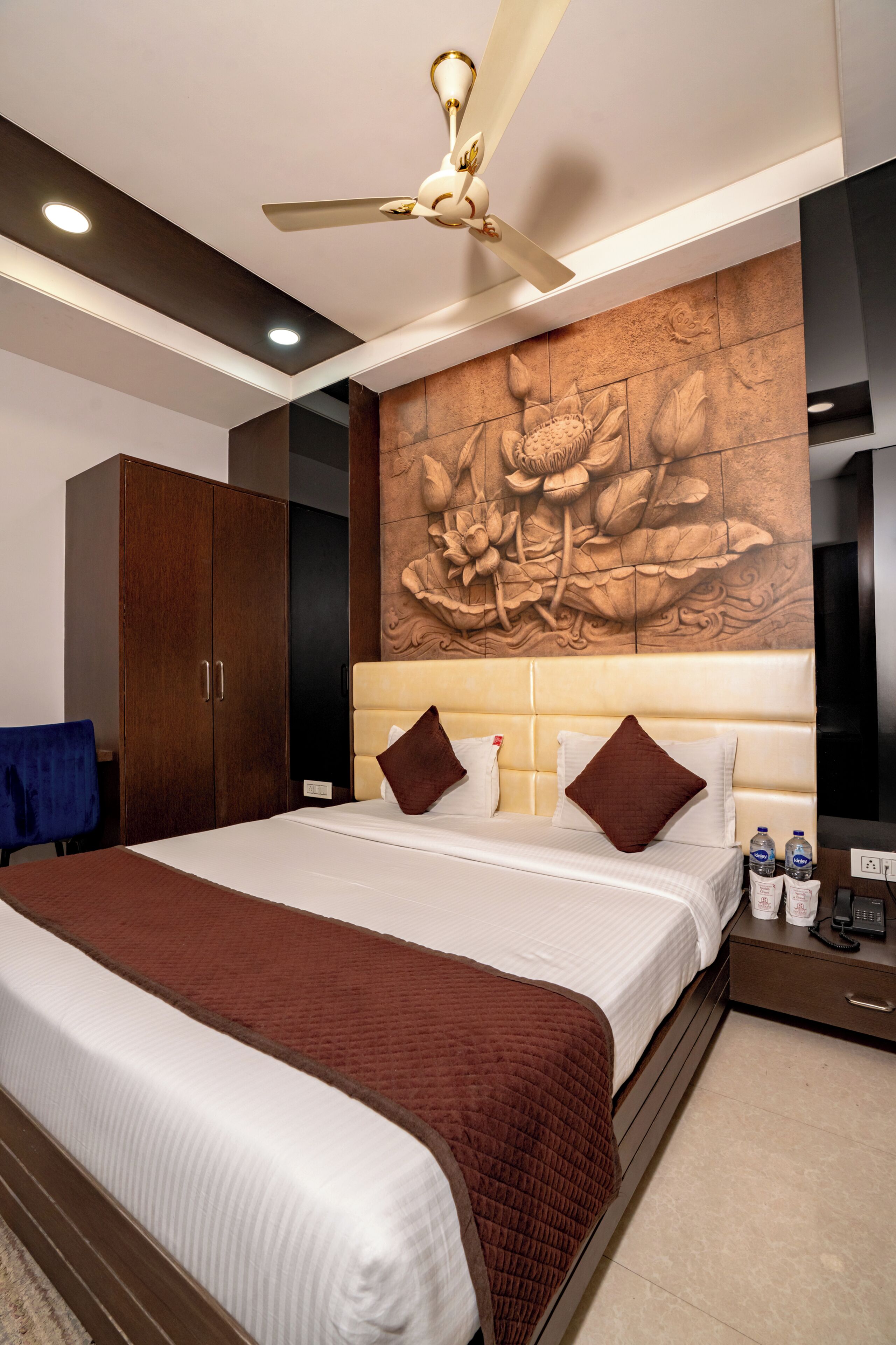 Quarto de hotel moderno com uma cama grande, ventoinha no teto, obra de arte na parede, guarda-roupa e mesa de cabeceira com telefone e garrafas de água.