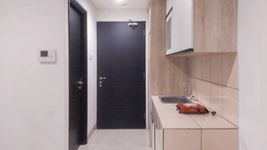 Um corredor estreito com uma porta preta no final, um pequeno painel de controle na parede e uma pia com um objeto vermelho no balcão.