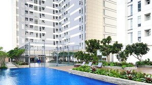 Piscina com deck de madeira, cercada por vegetação e um edifício alto ao fundo.