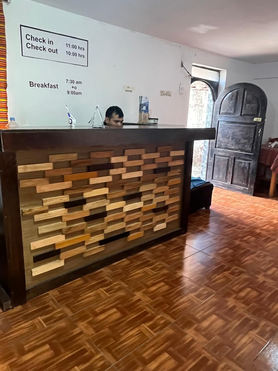 Bancada de recepção com placa indicando check-in/check-out, horários do café da manhã e piso de madeira.