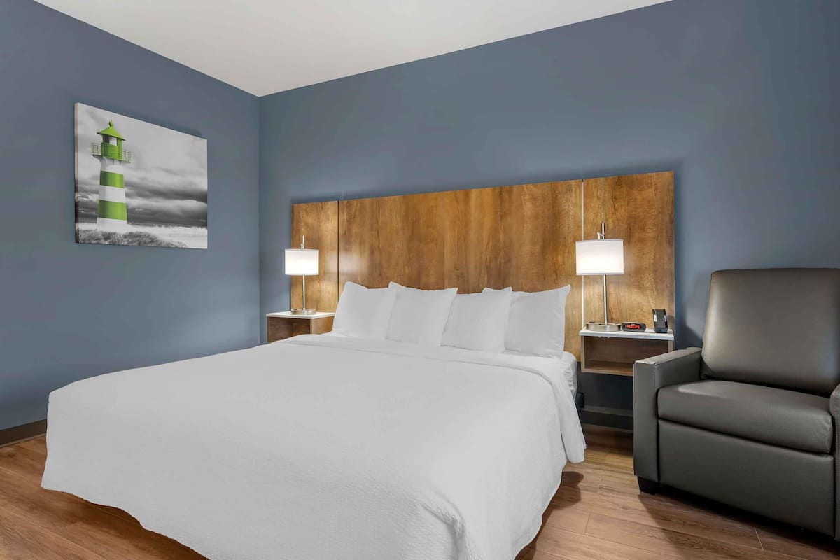 Quarto de hotel com uma cama grande, duas mesas de cabeceira com luminárias, um painel de madeira na cabeceira e uma poltrona cinza.
