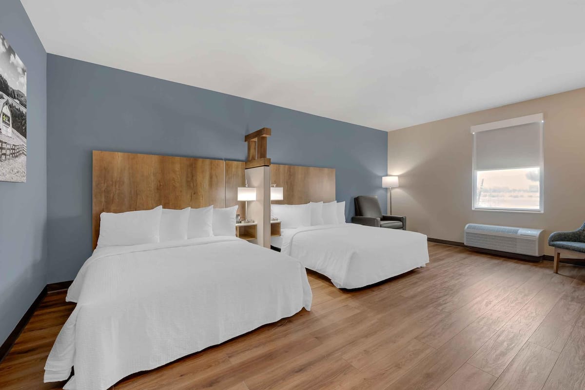 Quarto de hotel com duas camas, cabeceira de madeira, mesa de cabeceira, uma cadeira, janela com vista e ar condicionado fixado na parede.