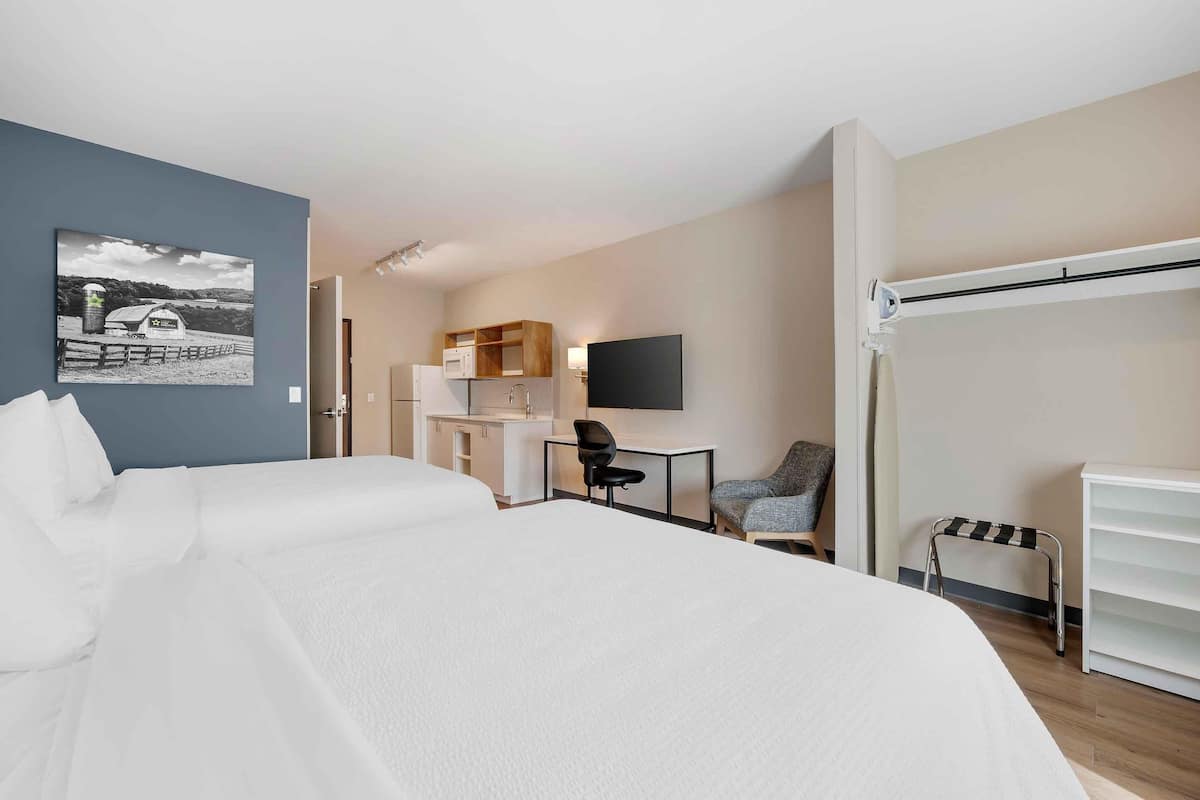 Quarto de hotel com uma cama grande, uma mesa com cadeira, televisão, guarda-roupa e um banquinho.