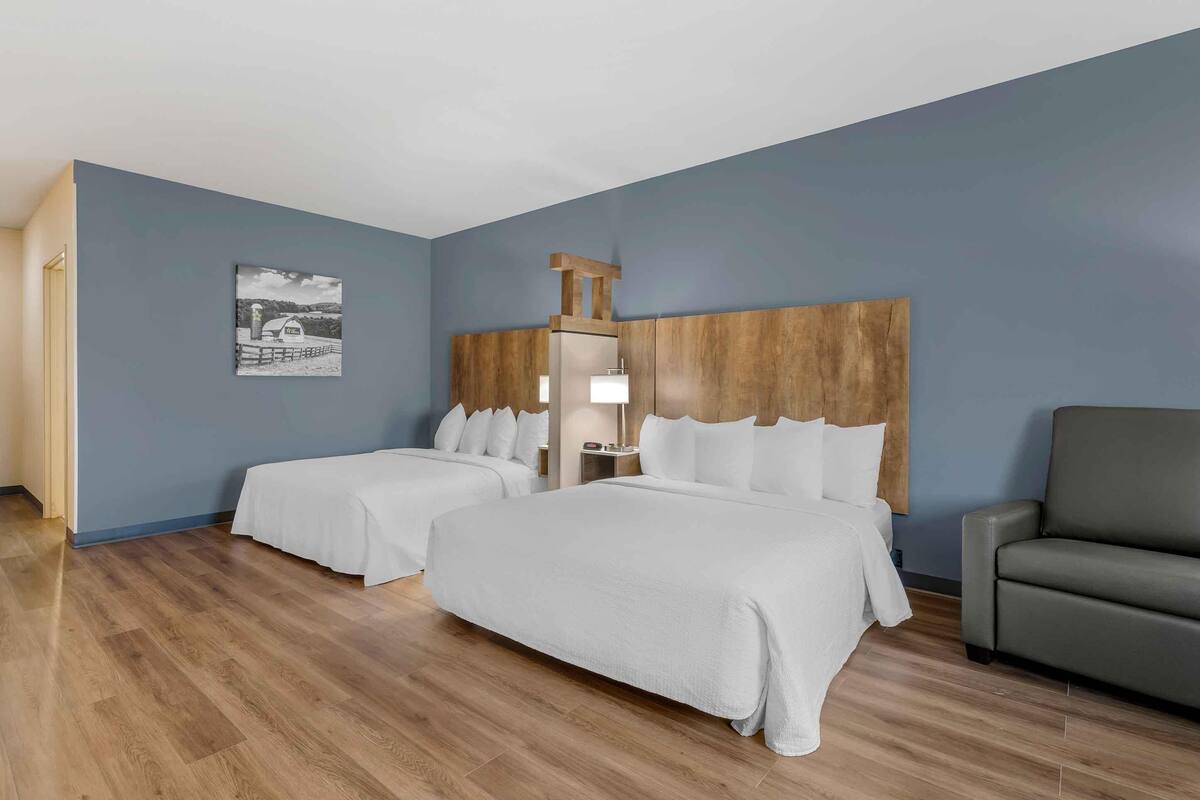 Quarto de hotel com duas camas, cabeceira de madeira, uma poltrona cinza e mesa de cabeceira com abajur.