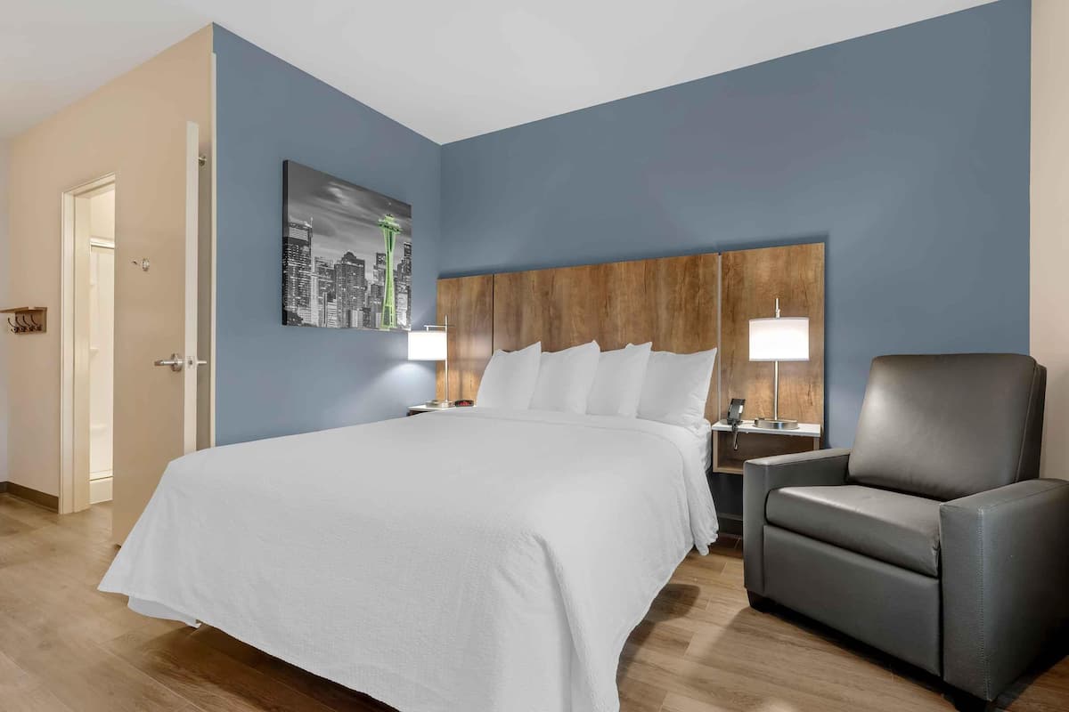 Quarto de hotel com uma cama grande, cabeceira de madeira, duas mesas de cabeceira com luminárias e uma poltrona de couro preto.