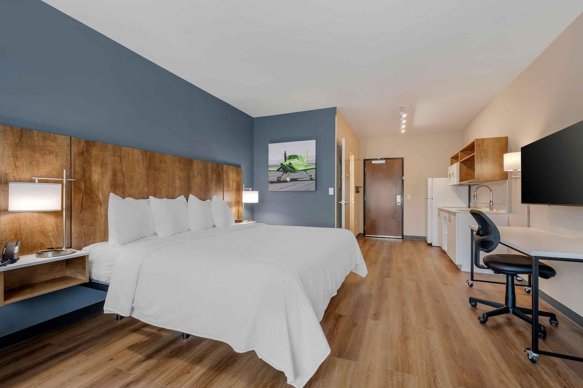 Quarto de hotel com cama, mesa de cabeceira, luminária, escrivaninha com cadeira e um quadro na parede.