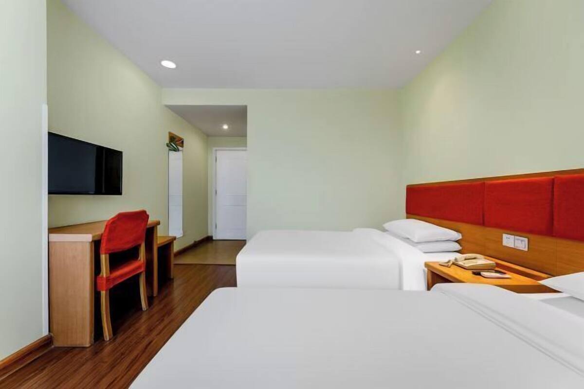 Quarto de hotel com duas camas, uma escrivaninha com cadeira, televisão e cabeceira vermelha.