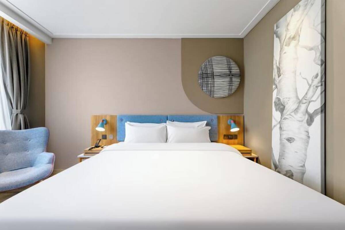 Quarto de hotel com cama, cabeceira azul, duas luminárias de apoio, uma cadeira e um painel na parede com uma obra de arte circular.