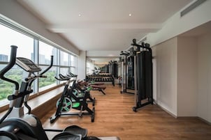 Academia com equipamentos de cardio, aparelhos de musculação e amplas janelas com vista para a cidade.