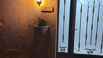 Um interior rústico com parede texturizada, um banco de madeira, uma planta em vaso, luminária de parede e porta de vidro decorativa com grade de metal.