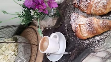 Um cenário de café da manhã com croissants polvilhados com açúcar de confeiteiro, uma xícara de café e um vaso decorativo com flores roxas.