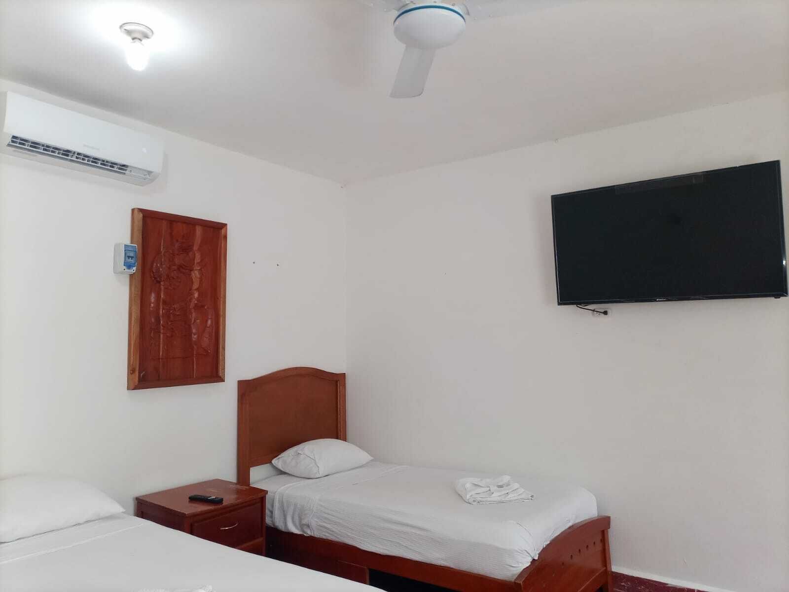 Quarto individual com uma cama e cabeceira de madeira, mesa de cabeceira, televisão fixada na parede e ar condicionado.