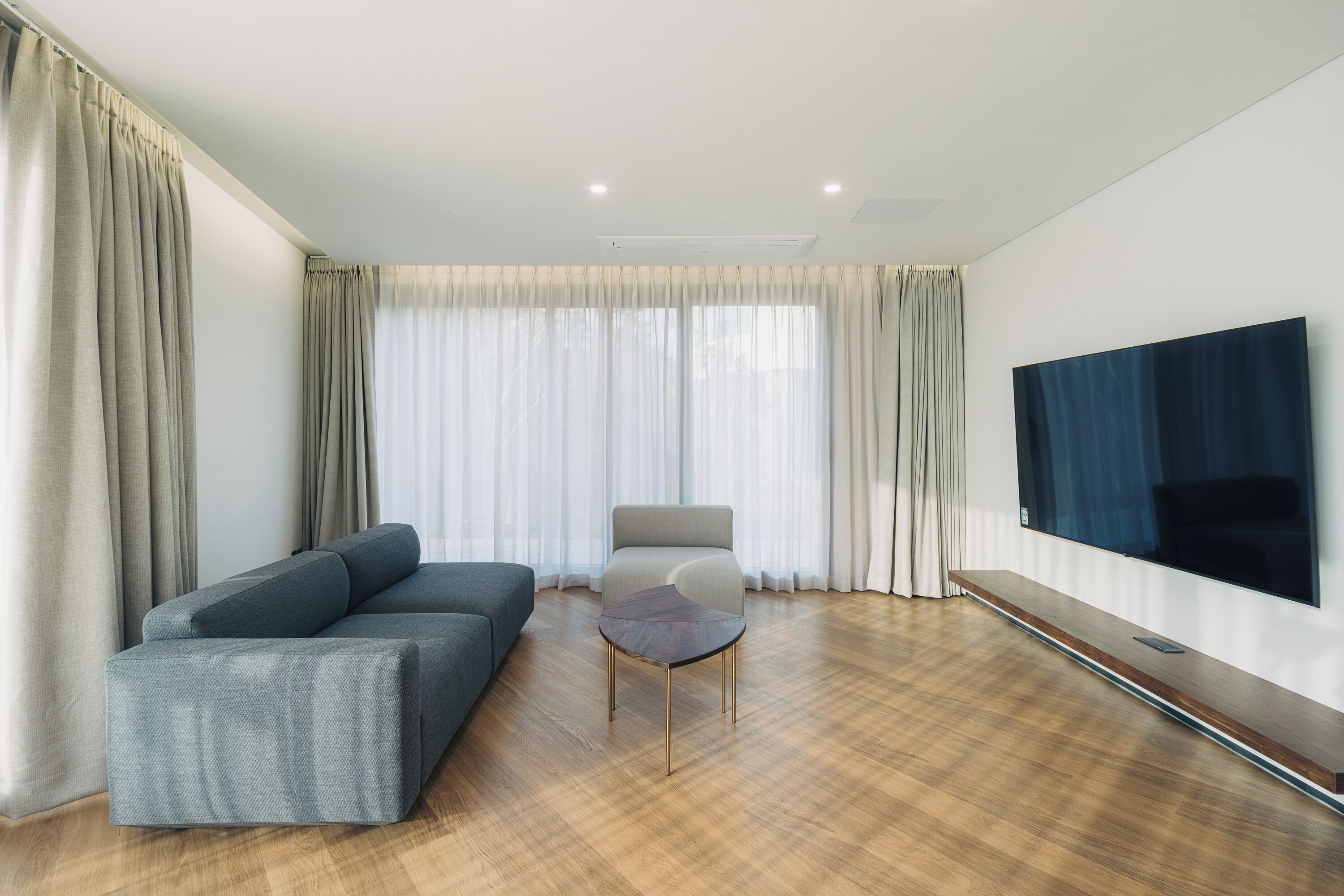 Uma sala de estar moderna com sofá, poltrona e mesa de centro, janelas amplas com cortinas e uma televisão de tela plana na parede.