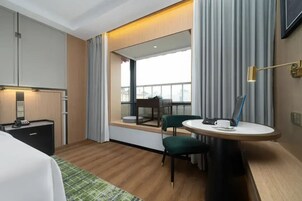 Quarto de hotel com uma cama, uma mesa com um laptop, uma cadeira, uma mesinha e uma janela com cortinas.