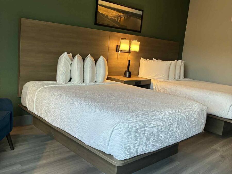 Uma cama de casal com roupa de cama branca, uma mesa de cabeceira com abajur e um quadro acima da cama.