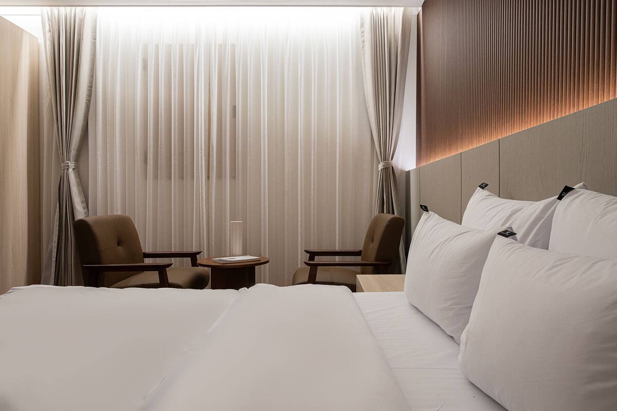Quarto de hotel com uma cama grande, duas poltronas, uma mesinha e cabeceira com linhas verticais.