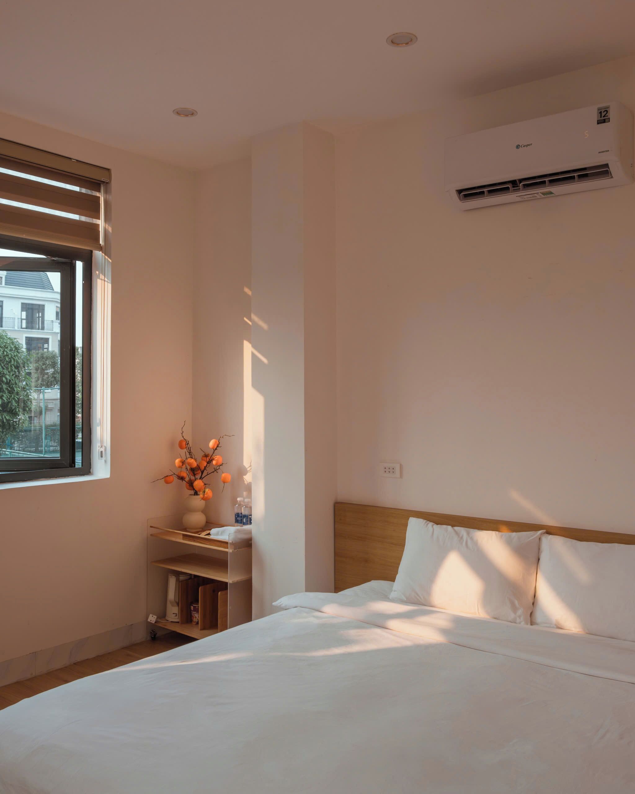 Quarto de hotel com cama, mesa de cabeceira com um vaso de flores, janela com persianas e um aparelho de ar condicionado na parede.