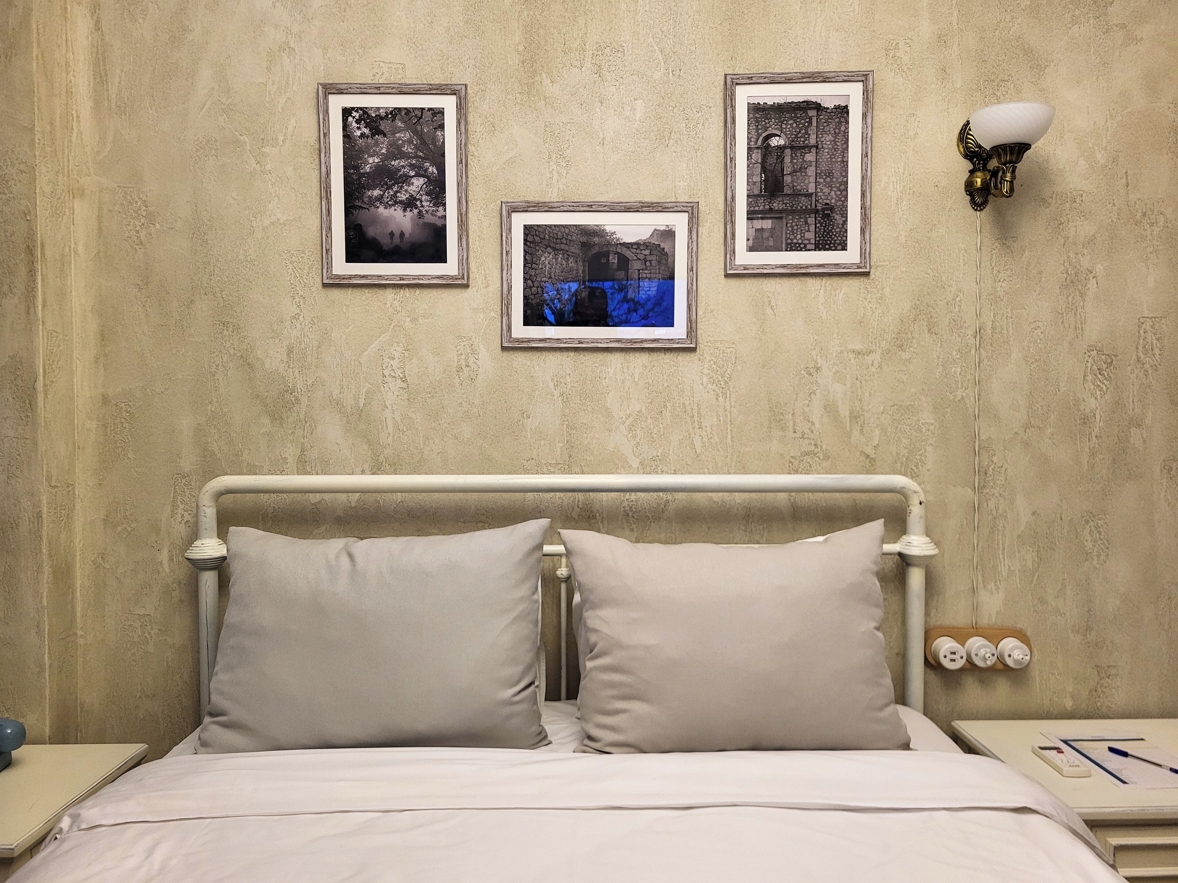Quarto com cama, duas almofadas, mesa de cabeceira com abajur e três quadros na parede.