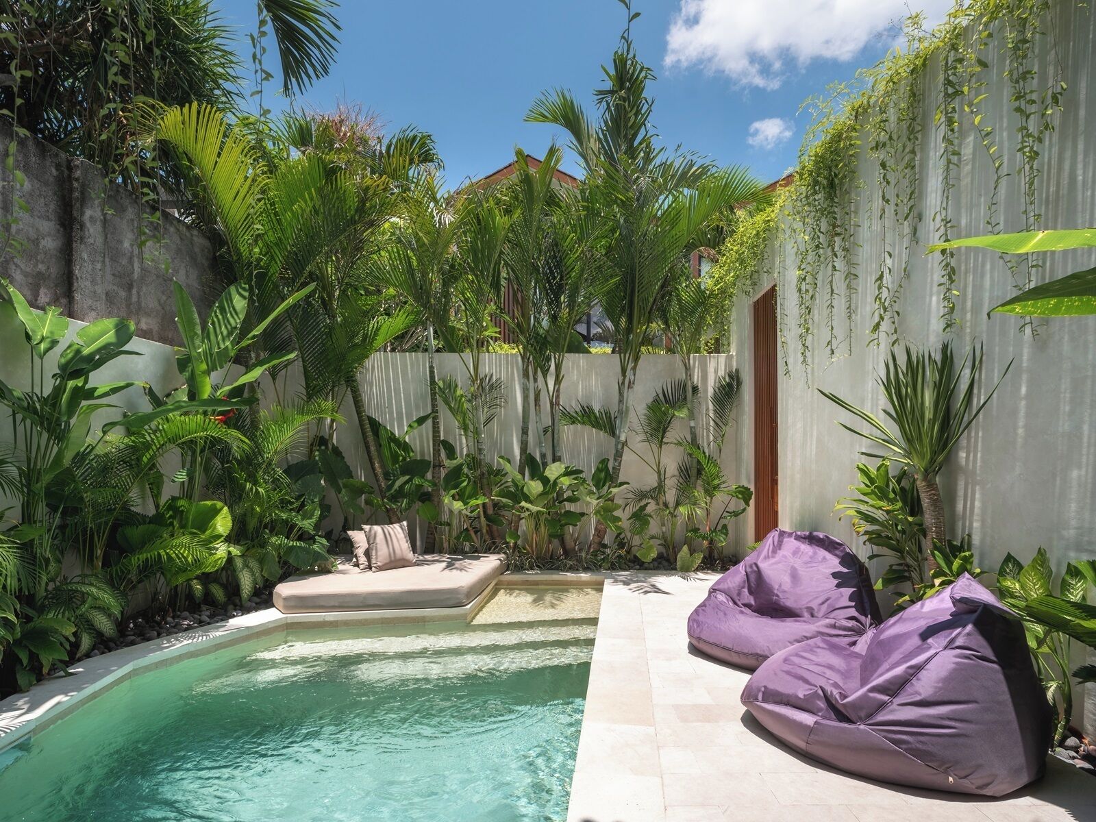 Uma área de piscina com borda de concreto, cercada por plantas tropicais exuberantes e um cercado de madeira.