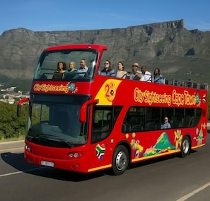 Um autocarro vermelho de dois andares com a inscrição 'CitySightseeing Cape Town' na lateral, passageiros visíveis e uma paisagem montanhosa ao fundo.