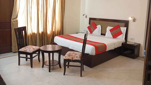Quarto de hotel com cama, duas cadeiras, uma mesinha, uma mesa de cabeceira e cortina.