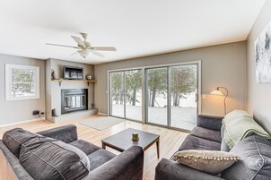 Uma sala de estar moderna com lareira, ventilador de teto e uma grande porta de vidro deslizante que dá para uma área externa coberta de neve.