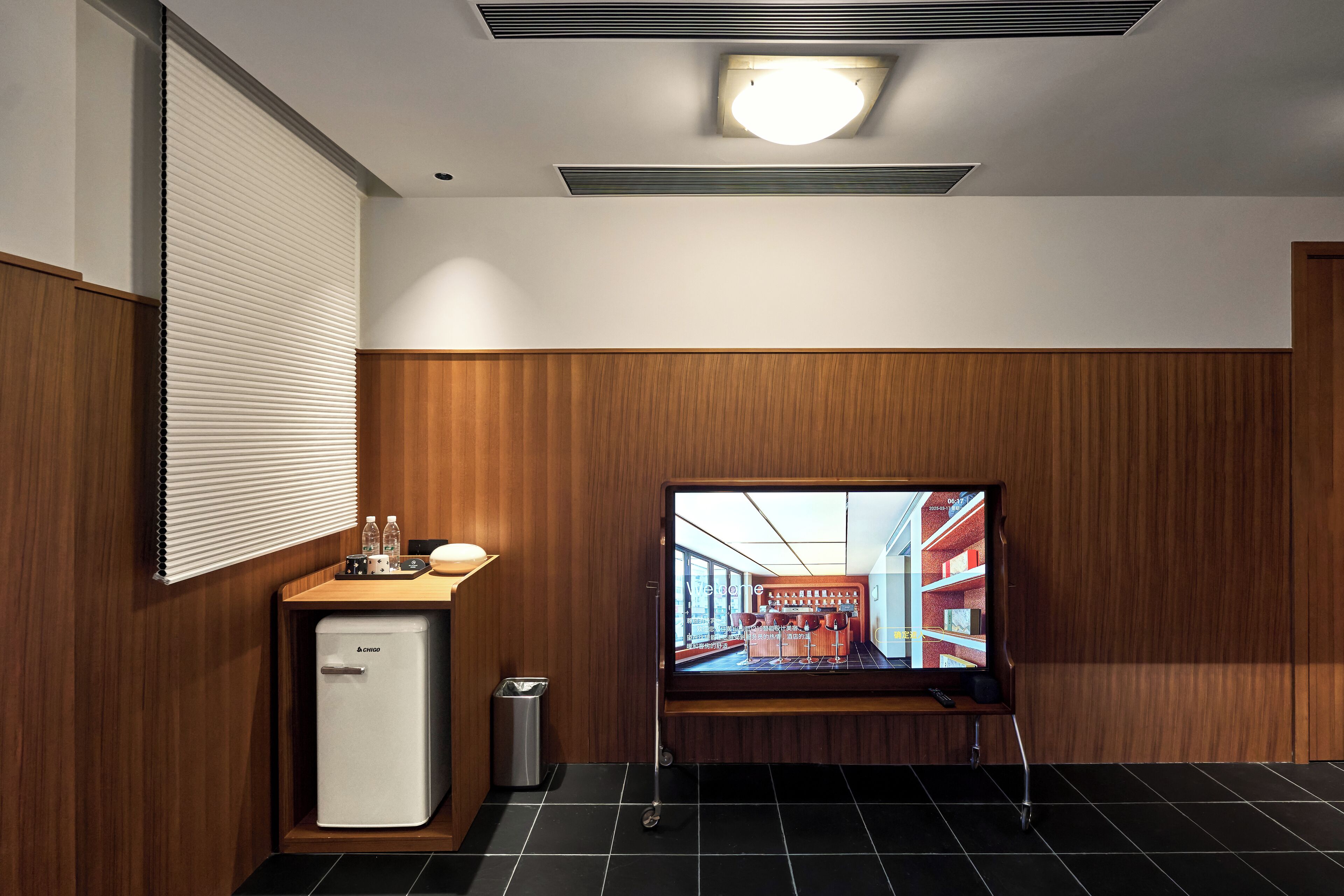 Quarto de hotel com parede revestida de madeira, pequena geladeira, lixeira e televisão sobre um suporte de madeira.