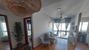 Uma sala de estar ampla com sofá, mesa de jantar e cadeiras. Há uma janela grande com cortinas, um ventilador de teto e um vaso de planta.