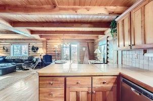 Interior de cabana de tronco com área de jantar de madeira, cozinha e sala de estar com sofá e cadeiras.