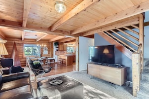 Interior espaçoso de uma cabana de madeira com área de jantar, sala de estar com sofá de couro, televisão e escada.