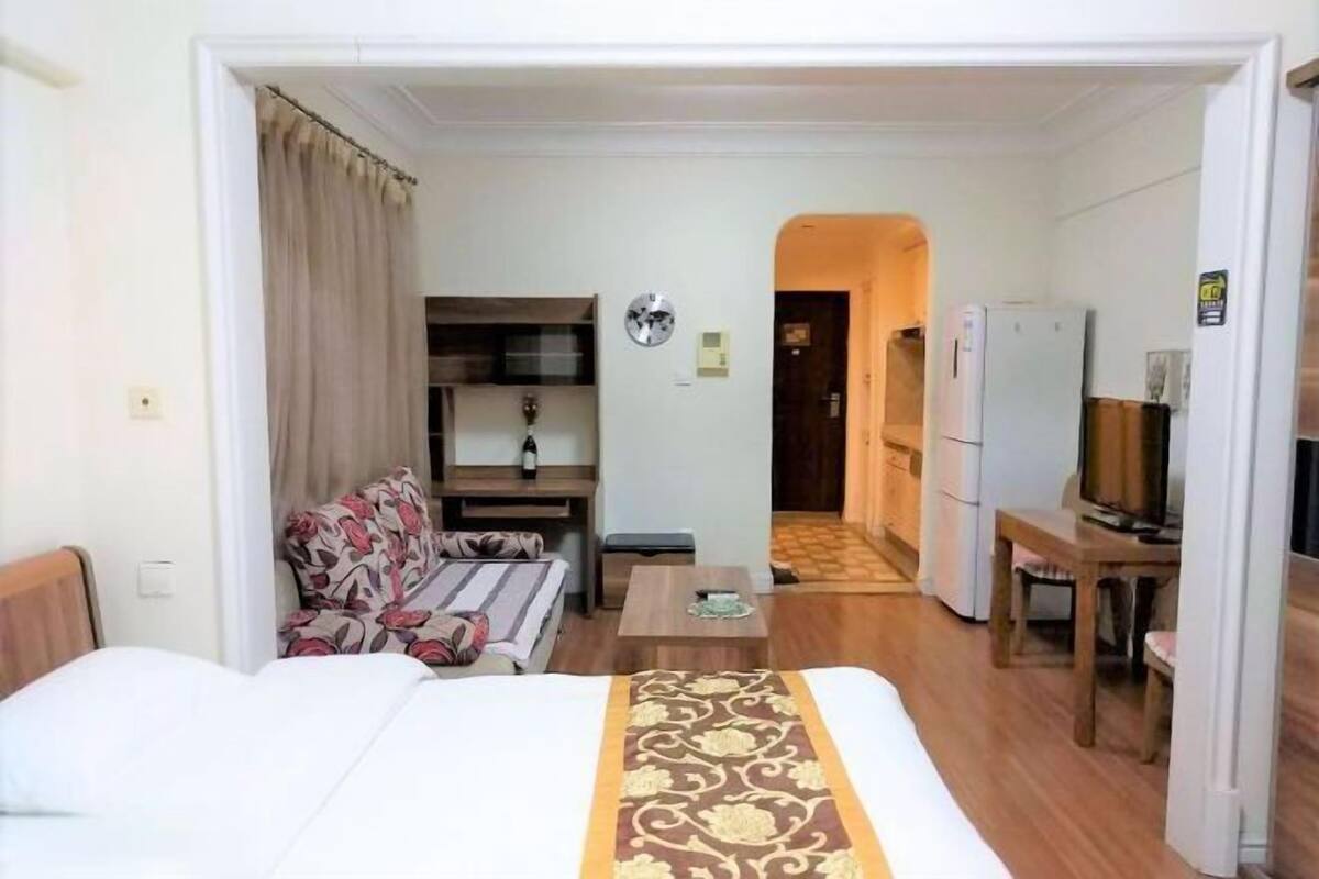 Quarto de hotel com cama, sofá, mesa pequena, televisão e cozinha compacta.