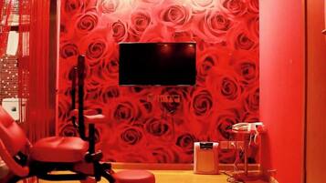 Quarto com papel de parede floral vermelho, televisão de tela plana na parede, uma cadeira vermelha, uma mesa pequena e um alto-falante.