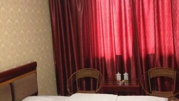 Quarto de hotel com uma cama, duas cadeiras de vime, uma mesinha de centro com abajur e cortinas vermelhas.