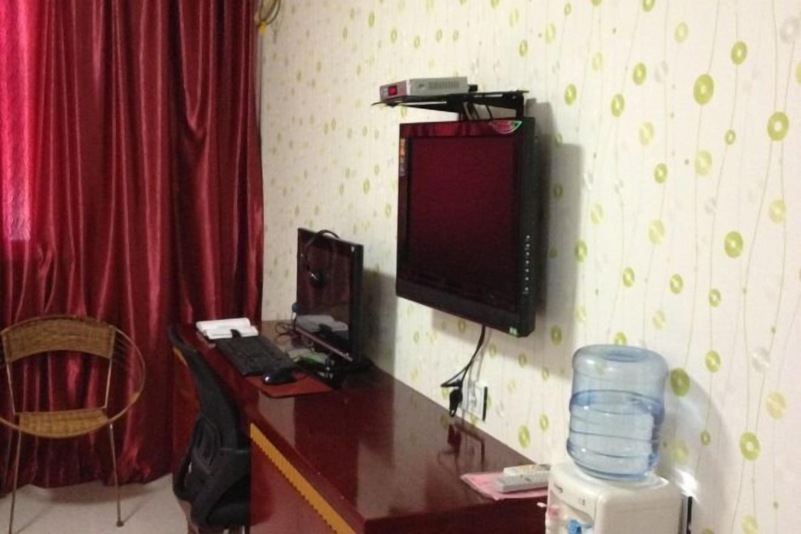 Quarto com escrivaninha, cadeira, dispensador de água e televisão na parede.