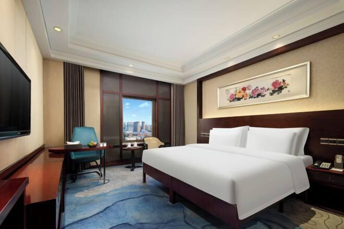 Quarto de hotel com uma cama grande, uma escrivaninha com cadeira, televisão e janela com vista para a cidade.