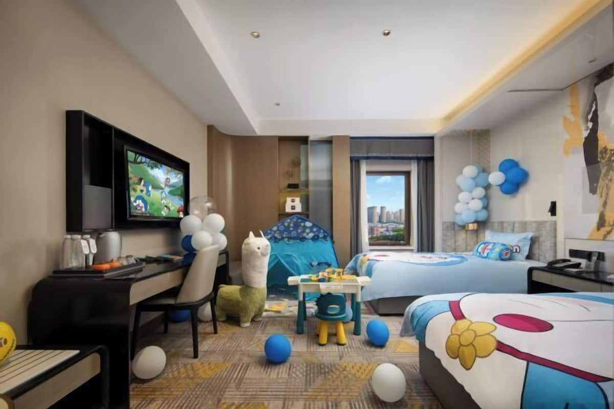 Quarto de hotel com televisão, escrivaninha, cadeira, cama, mesinha com brinquedos e janela com vista para a cidade.