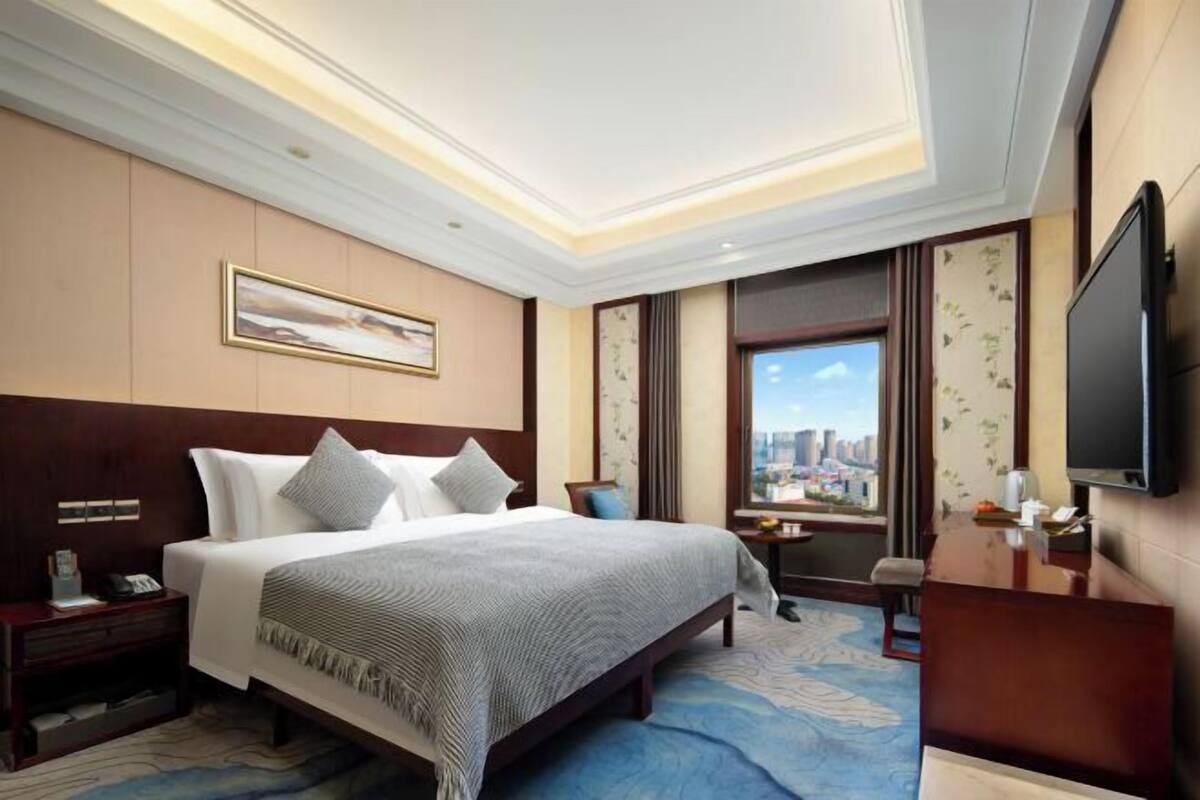 Quarto de hotel com uma cama grande, uma escrivaninha, uma televisão e uma janela com vista para a cidade.