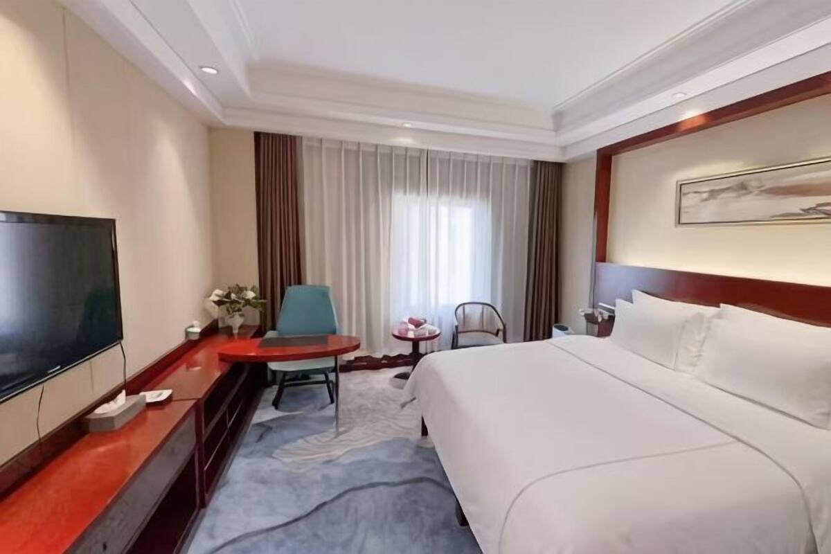 Quarto de hotel com cama, televisão, escrivaninha com cadeira e janela com cortinas.