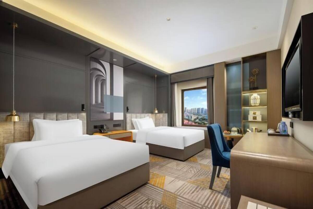 Quarto de hotel com duas camas, uma escrivaninha, uma cadeira, televisão e uma janela grande com vista para a cidade.