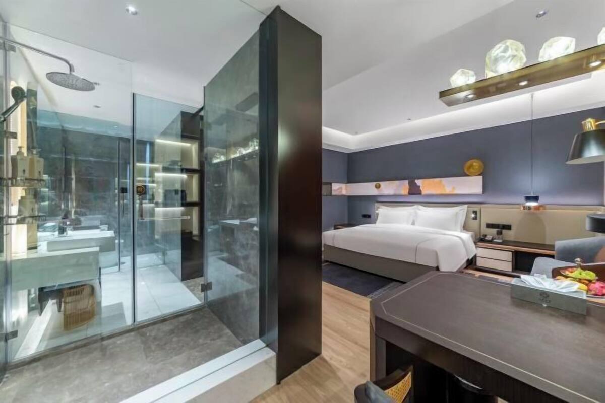 Quarto de hotel moderno com box de vidro, cama com roupa de cama branca, mesa de cabeceira e escrivaninha com cadeira.