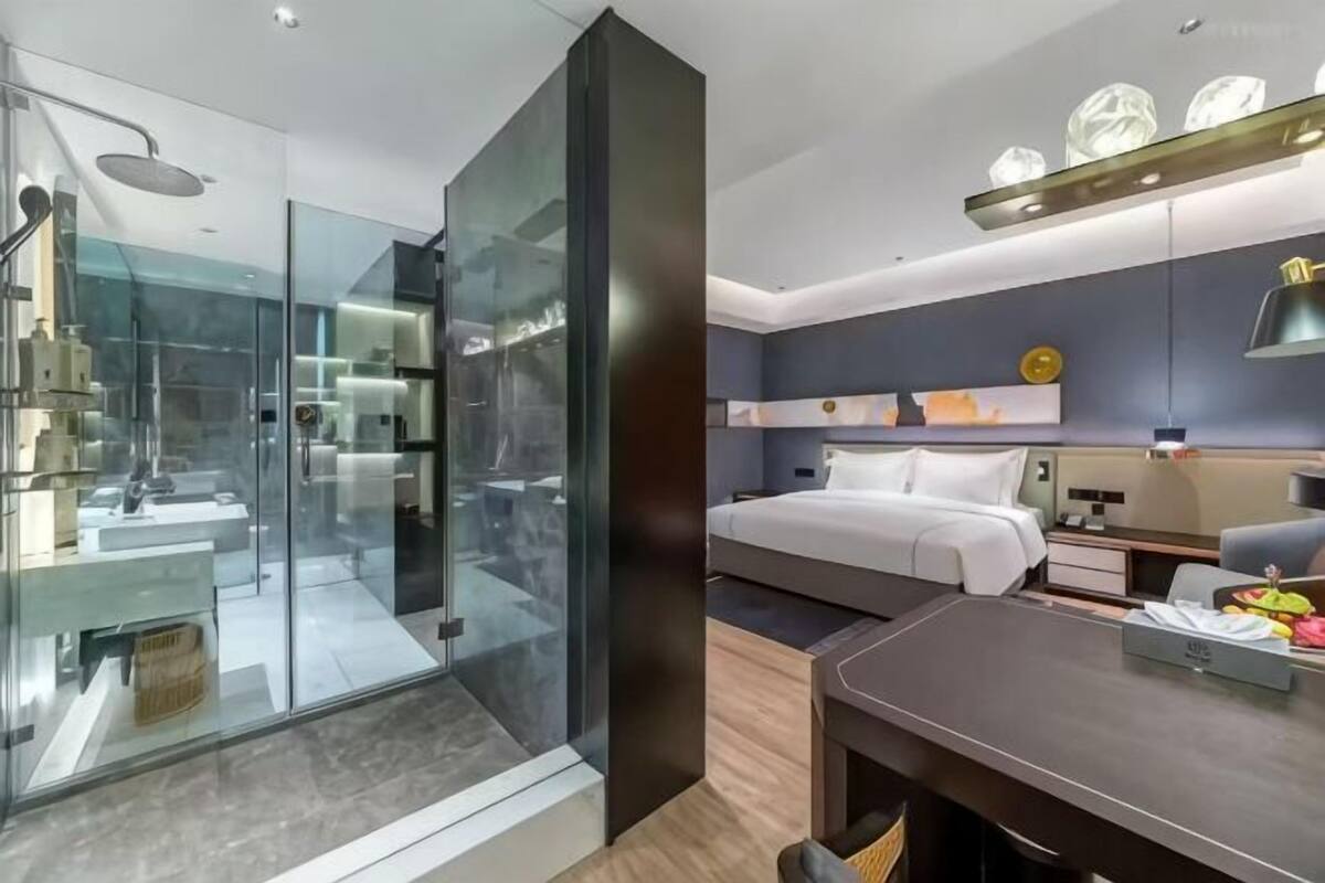 Quarto de hotel moderno com chuveiro em cabine de vidro, cama com cabeceira de madeira, mesa de cabeceira com abajur e pequena escrivaninha com cadeira.