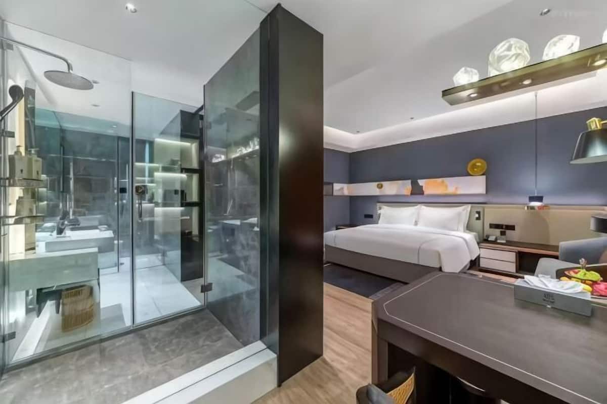 Quarto de hotel moderno com chuveiro em cabine de vidro, cama com cabeceira de madeira, mesa de cabeceira com abajur e pequena escrivaninha com cadeira.