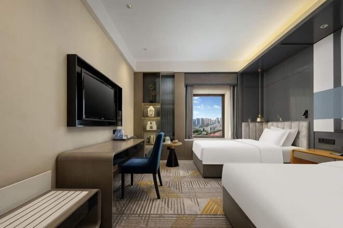 Quarto de hotel com cama, escrivaninha, cadeira, televisão e vista para a paisagem urbana.